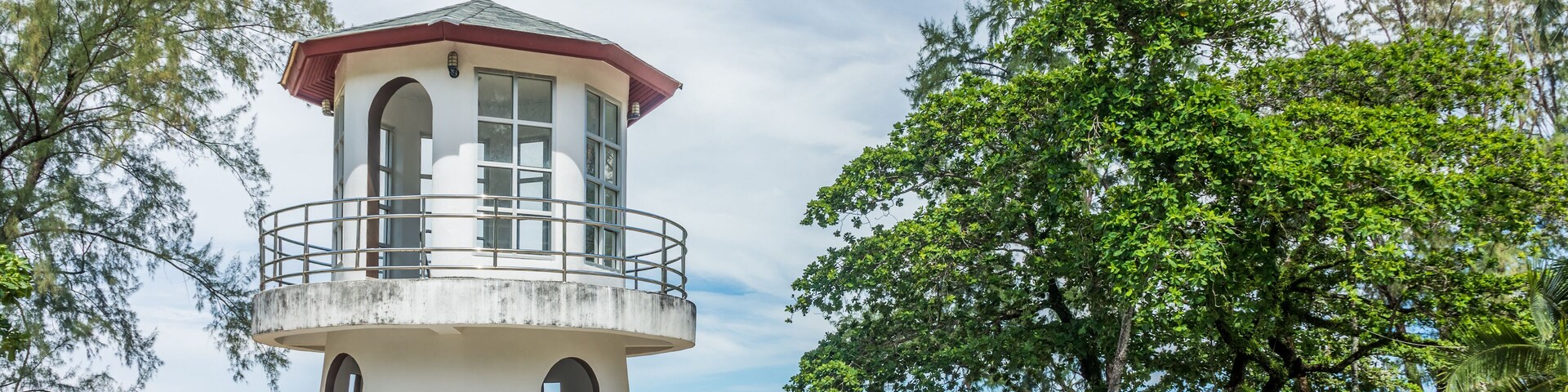 Um farol com varanda, localizado à beira-mar, rodeado por árvores e um céu claro.