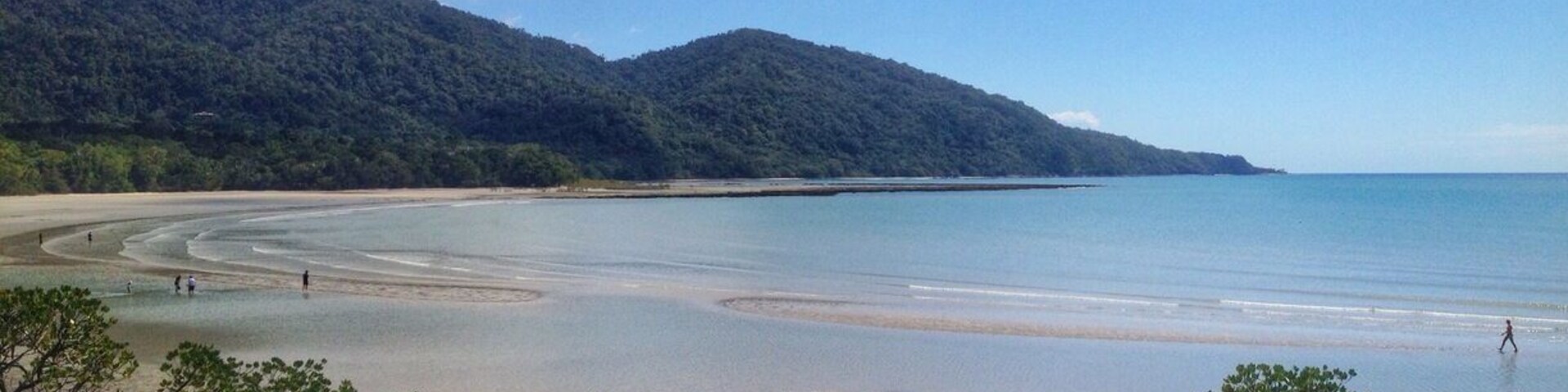 Uma praia com águas cristalinas, areal arenoso e colinas verdes ao fundo.