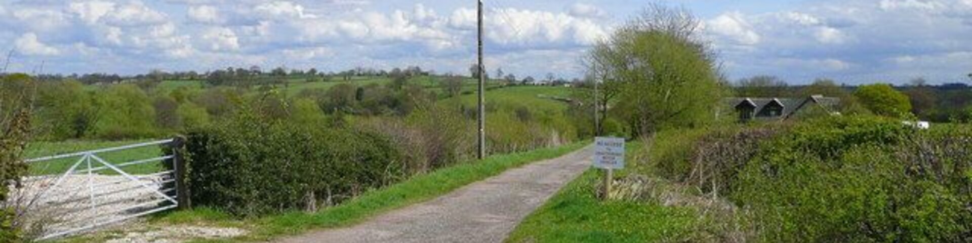 Uma estrada rural com um portão de metal, cercada por vegetação e um céu nublado.