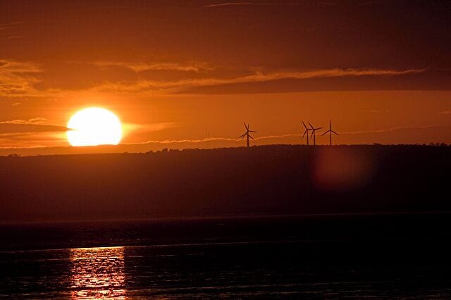 Um pôr do sol sobre a água, com turbinas eólicas recortadas no céu.