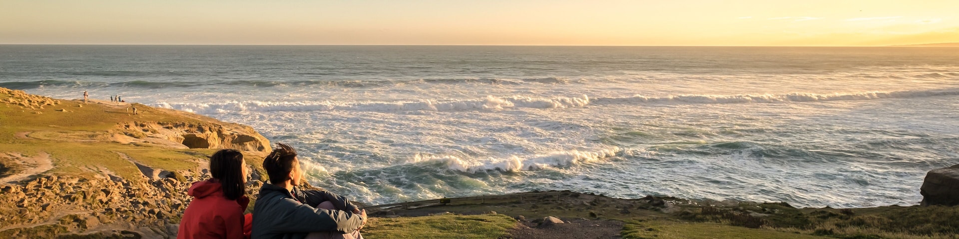 Um casal sentado num banco admirando o oceano ao pôr do sol.