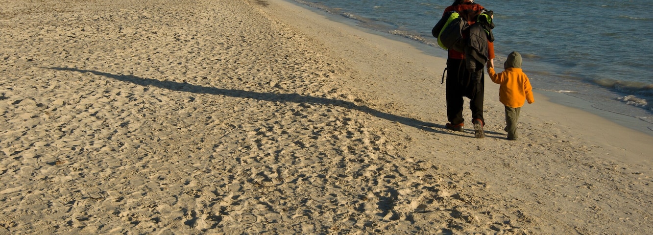 Uma praia arenosa com pegadas, uma pessoa carregando uma criança, e o oceano ao fundo.