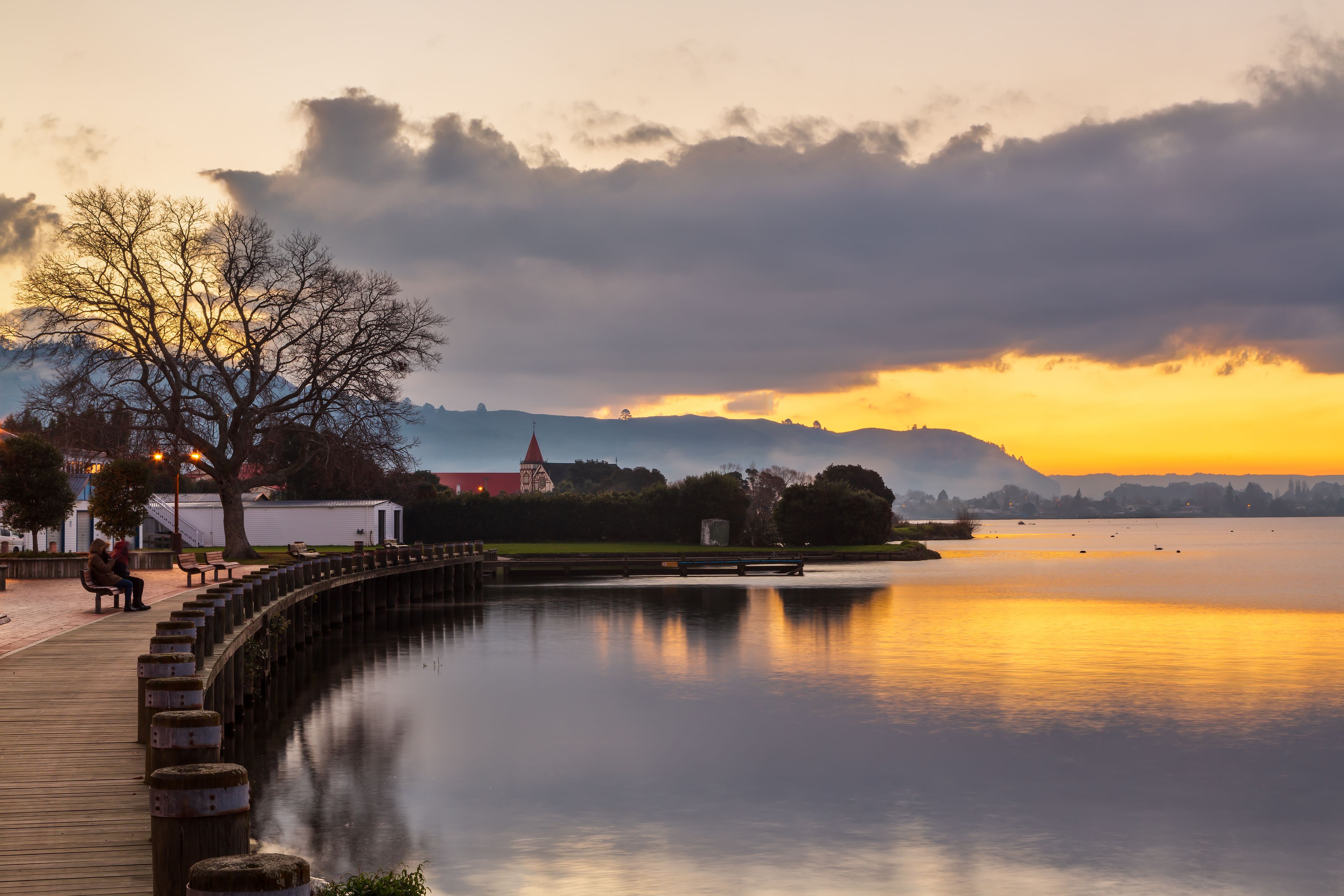 Um lago tranquilo ao pôr do sol, com uma passarela de madeira e bancos, um edifício com telhado vermelho e um caminho arborizado.