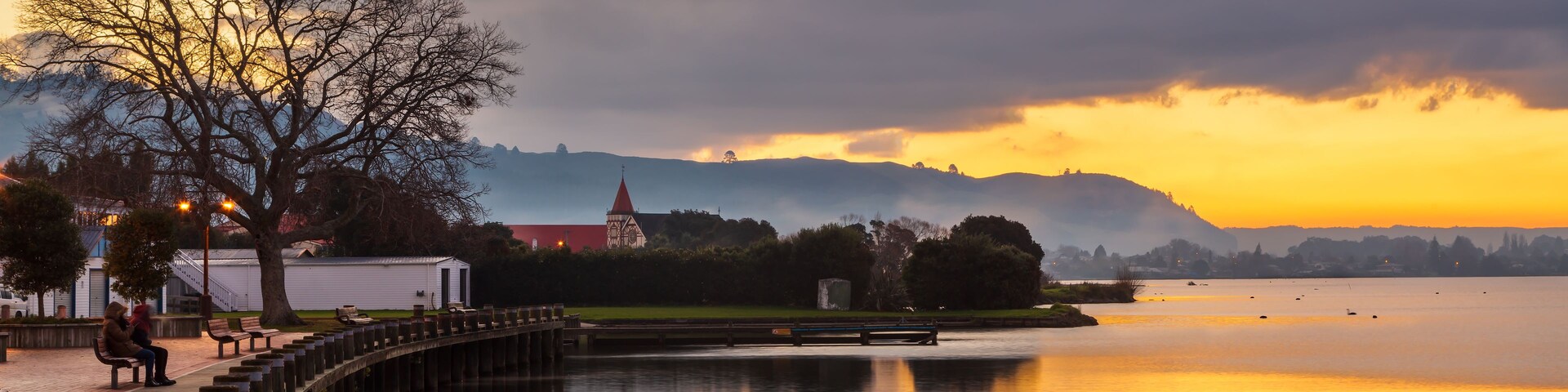 Um lago tranquilo ao pôr do sol, com uma passarela de madeira e bancos, um edifício com telhado vermelho e um caminho arborizado.