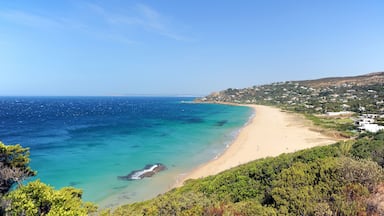 Paisagem costeira com praia arenosa, águas turquesa cristalinas e um relevo acidentado com edifícios dispersos.