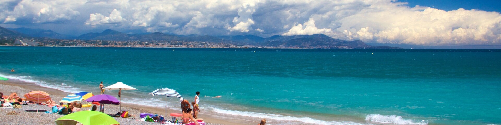 Uma praia de pedrinhas com pessoas se banhando e tomando sol, guarda-sóis coloridos e um mar azul cristalino sob um céu parcialmente nublado.