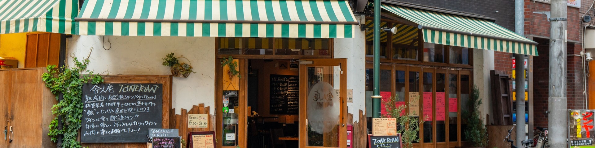 Fachada com toldo verde e branco listrado, uma bicicleta estacionada na frente e uma faixa de pedestres ao primeiro plano.