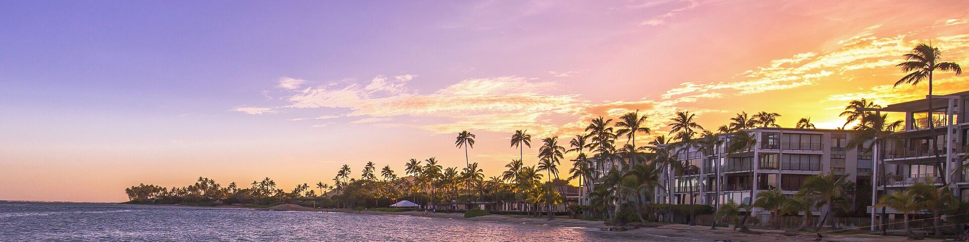 Um pôr do sol sobre uma praia com palmeiras e um hotel.