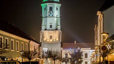 Um campanário com cúpula verde e luzes, rodeado por edifícios e decorações de Natal.