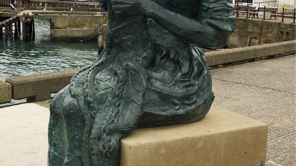Uma estátua de bronze de uma mulher sentada sobre um pedestal com peixes gravados, localizada em ambiente urbano com edifícios e um cais ao fundo.