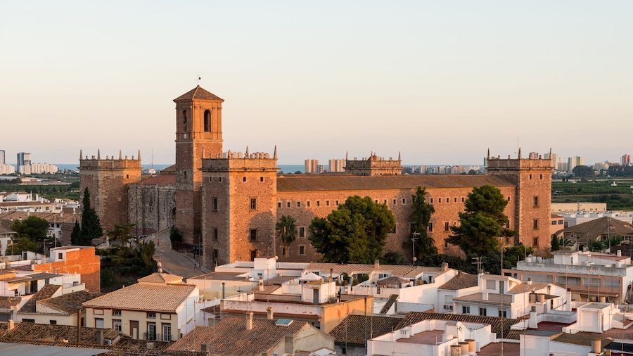 Uma fortaleza histórica com uma torre imponente, rodeada por edifícios residenciais e uma paisagem urbana ao fundo.