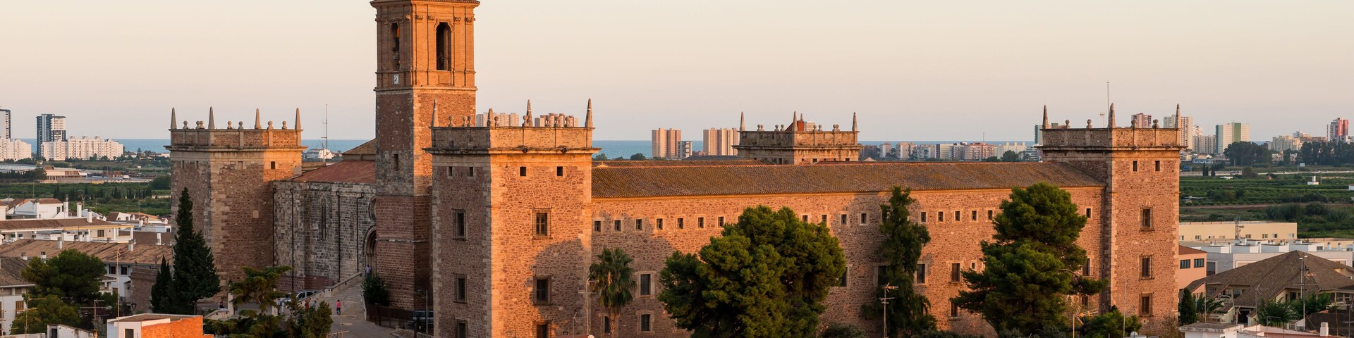 Uma fortaleza histórica com uma torre imponente, rodeada por edifícios residenciais e uma paisagem urbana ao fundo.