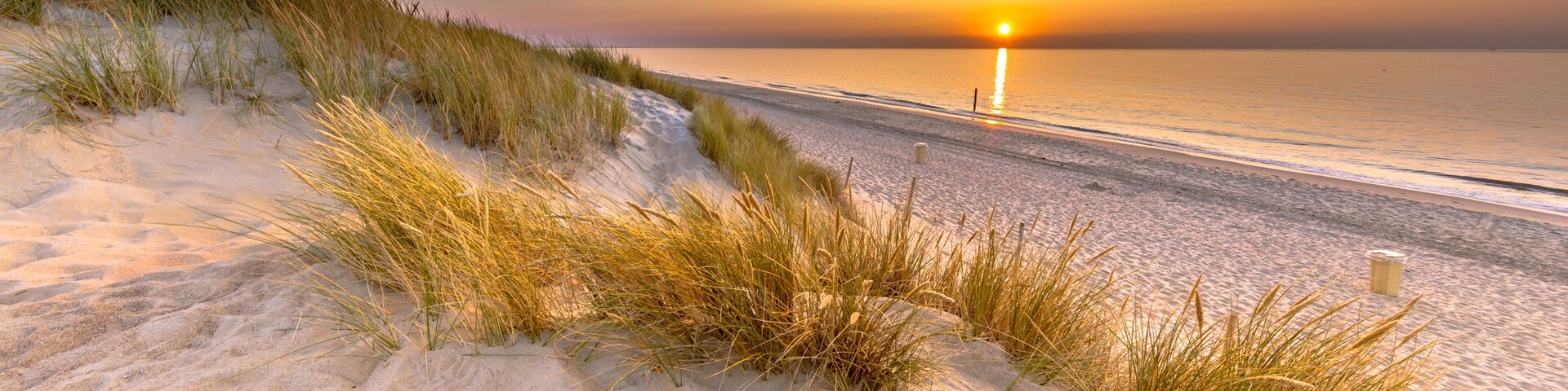 Uma praia ao pôr do sol, com dunas de areia e grama.