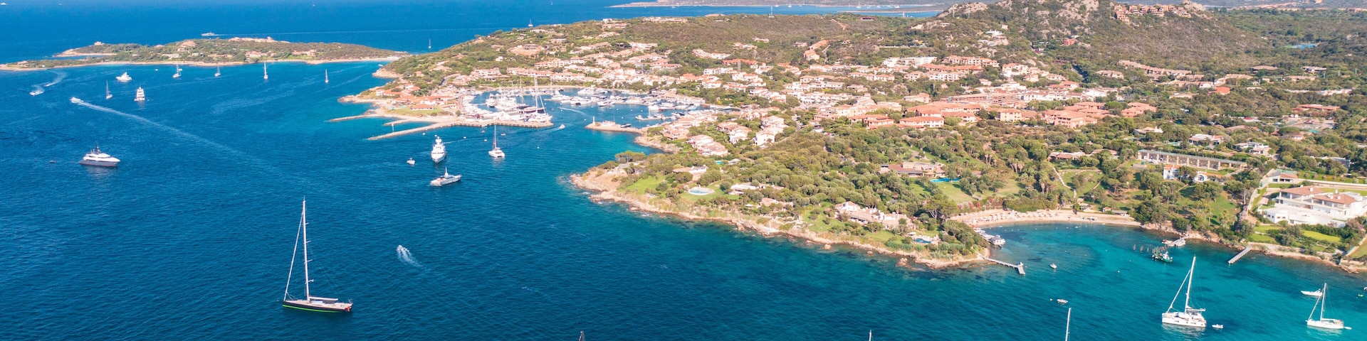 Uma vila costeira com veleiros na água, rodeada por colinas e mar azul cristalino.