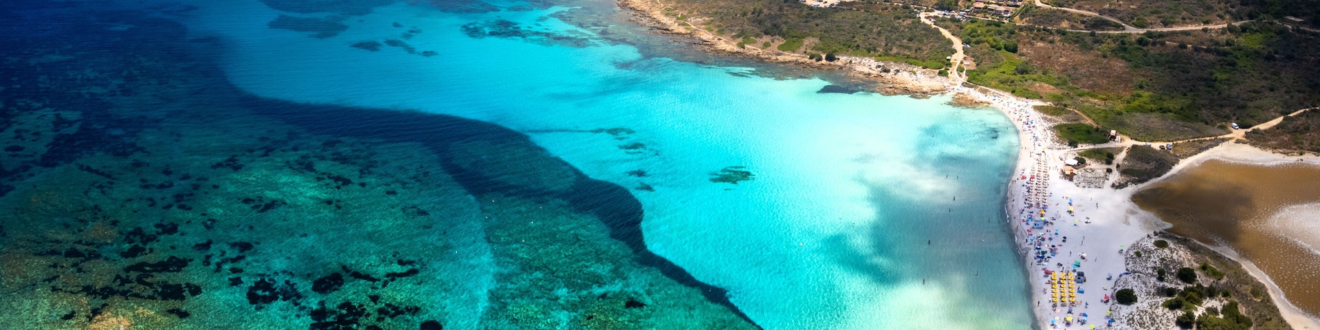 Paisagem costeira com águas turquesa cristalinas, uma praia arenosa e uma estrada sinuosa por uma área montanhosa.