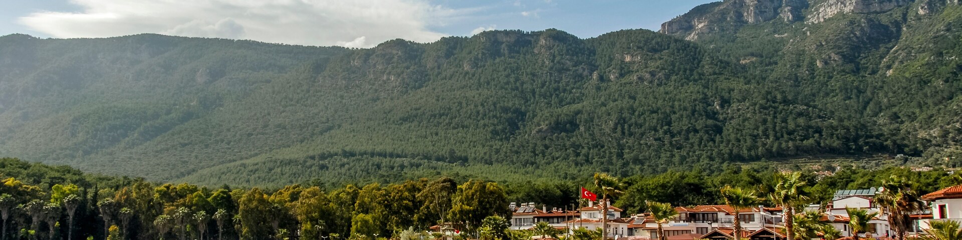 Uma praia costeira com montanhas ao fundo, céu claro e uma bandeira indicando localização turca.
