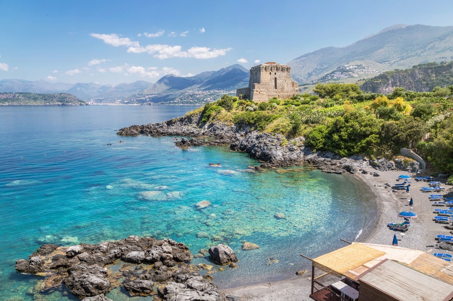 Paisagem costeira com águas turquesa cristalinas, uma linha costeira rochosa, uma torre histórica e uma praia com guarda-sóis.