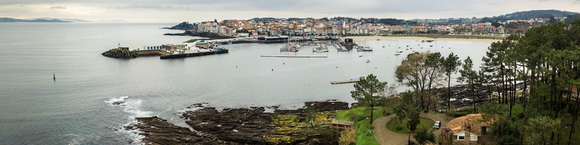 Uma cidade costeira com um porto de iates, costa rochosa e um cenário montanhoso ao fundo.