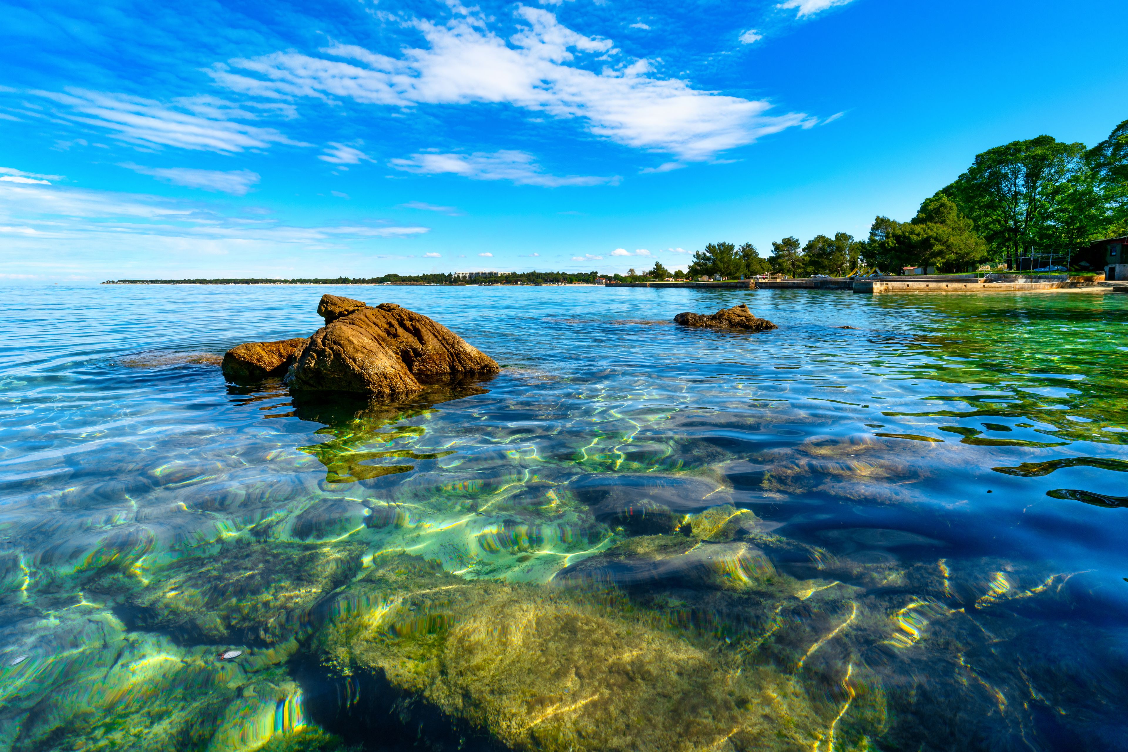 Lago calmo com pedras visíveis e uma linha de costa distante com edifícios e árvores.