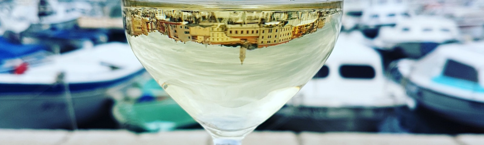 Um copo de vinho branco sobre uma mesa de madeira, com um porto e barcos ao fundo.