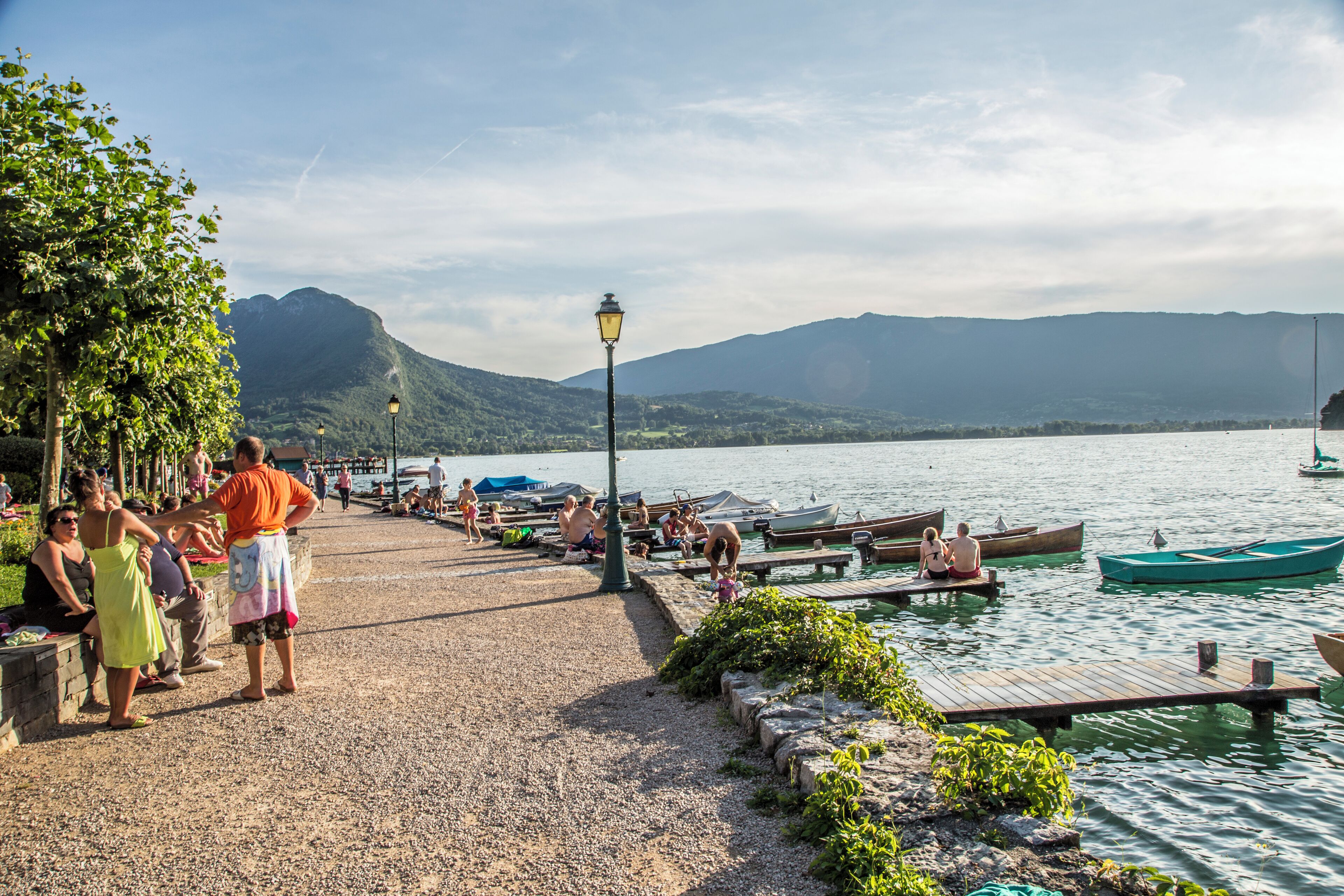 Um calçadão à beira do lago com pessoas relaxando, barcos atracados e montanhas ao fundo.