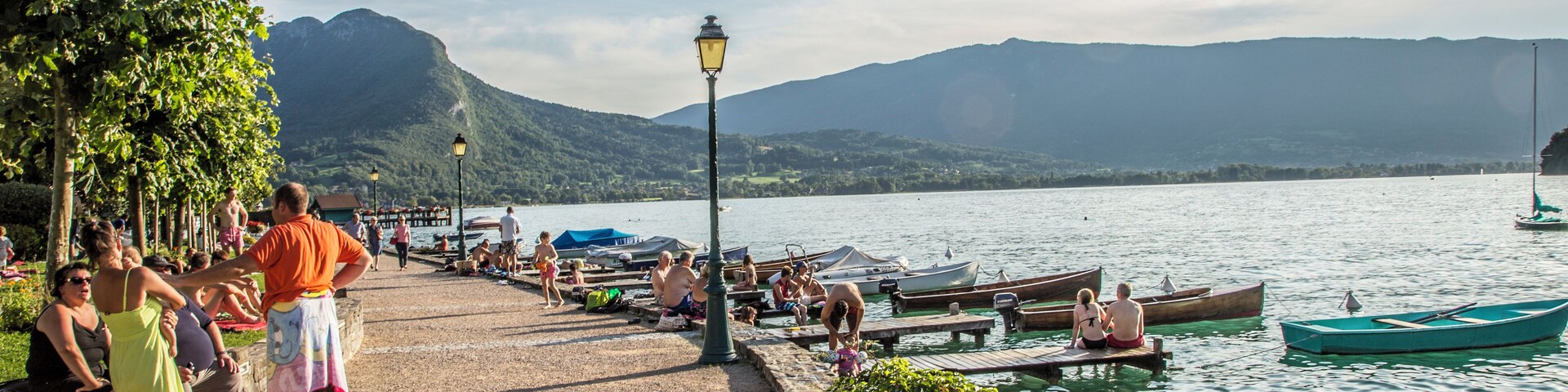 Um calçadão à beira do lago com pessoas relaxando, barcos atracados e montanhas ao fundo.