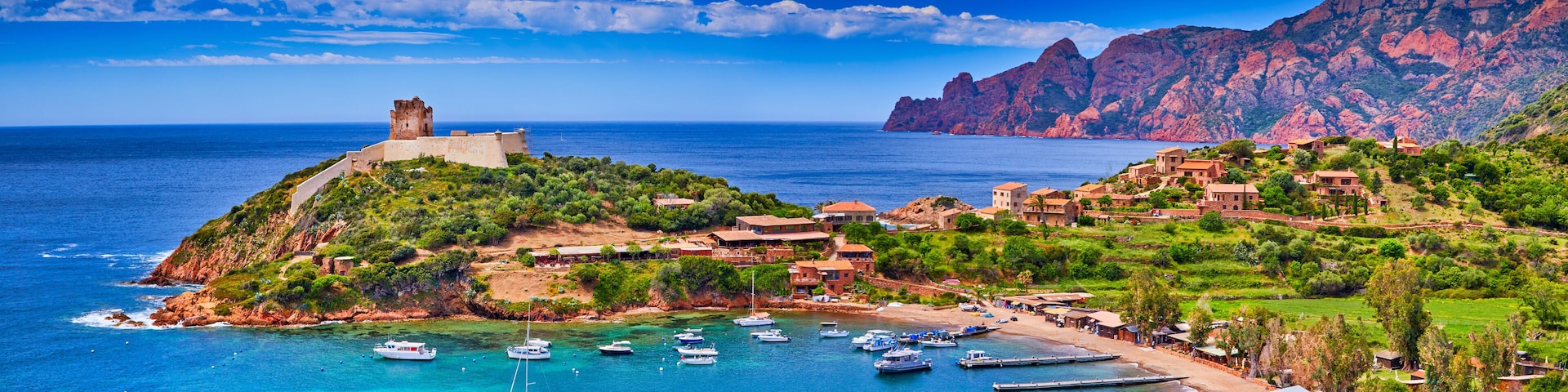 Uma cidade costeira com um castelo histórico, águas turquesa cristalinas e relevo montanhoso ao fundo.