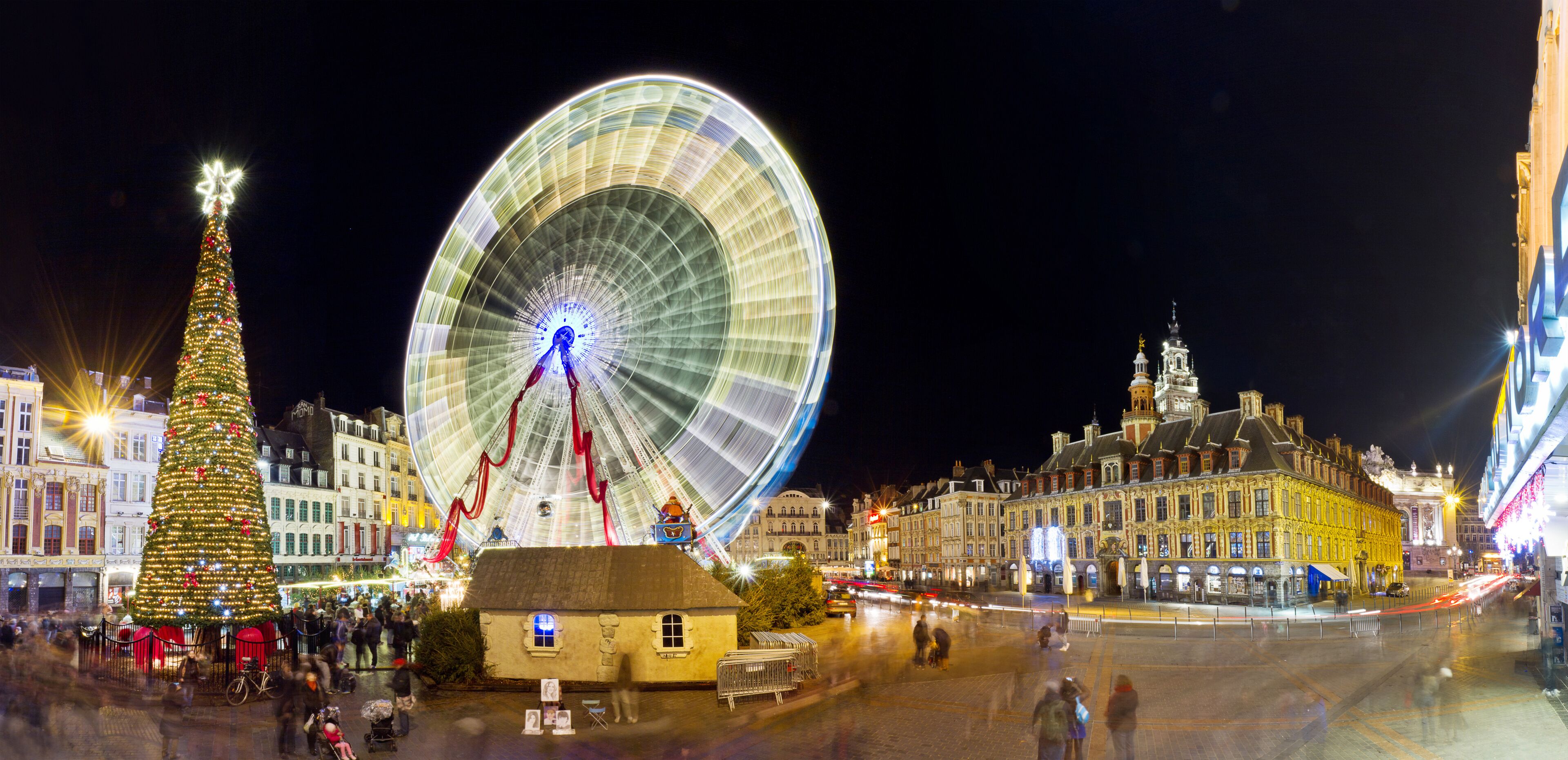 Uma árvore de Natal decorada, uma roda gigante e edifícios históricos em uma praça da cidade à noite.
