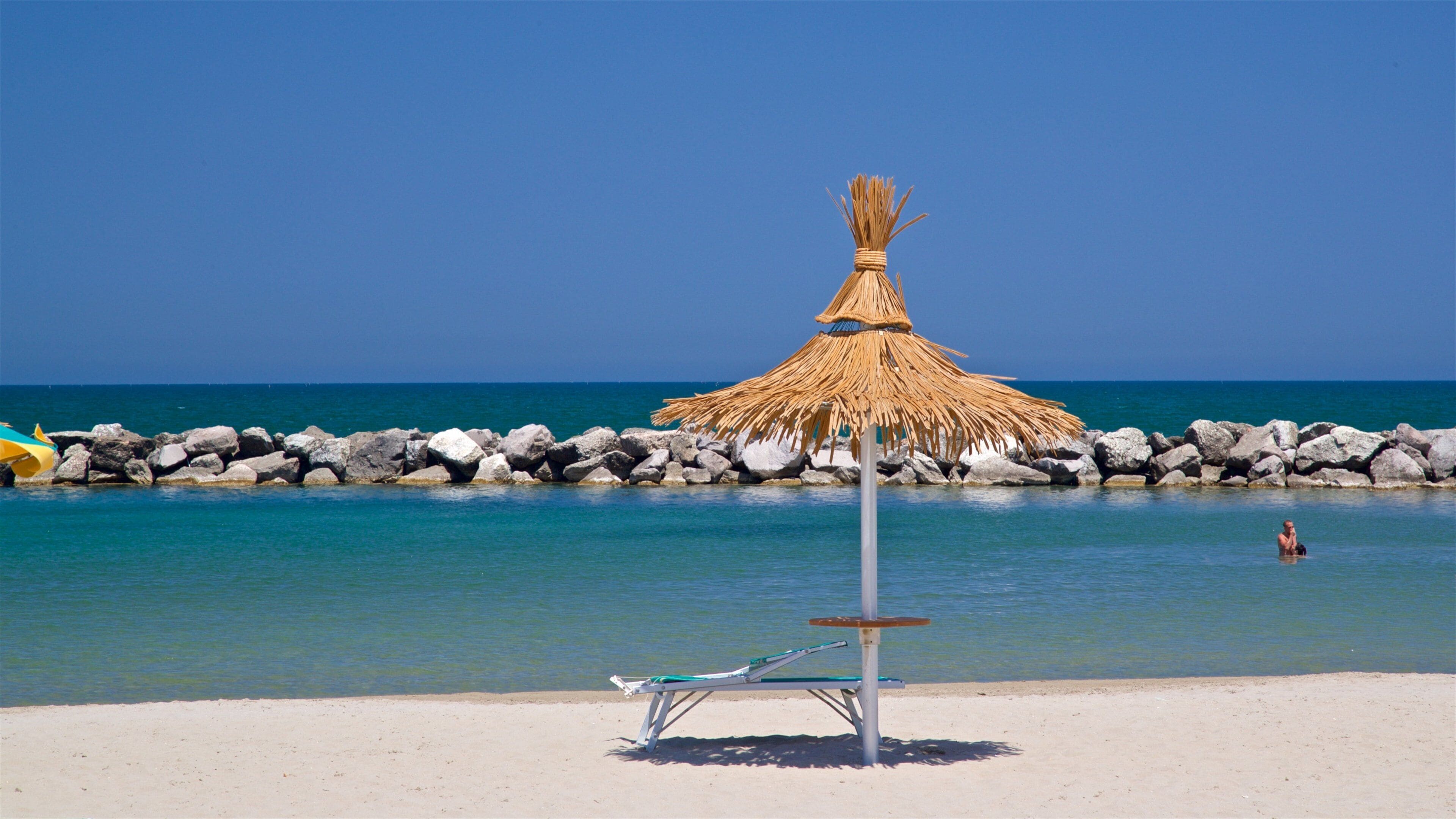 Uma praia com um guarda-sol de palha, um molhe rochoso e águas turquesa calmas.
