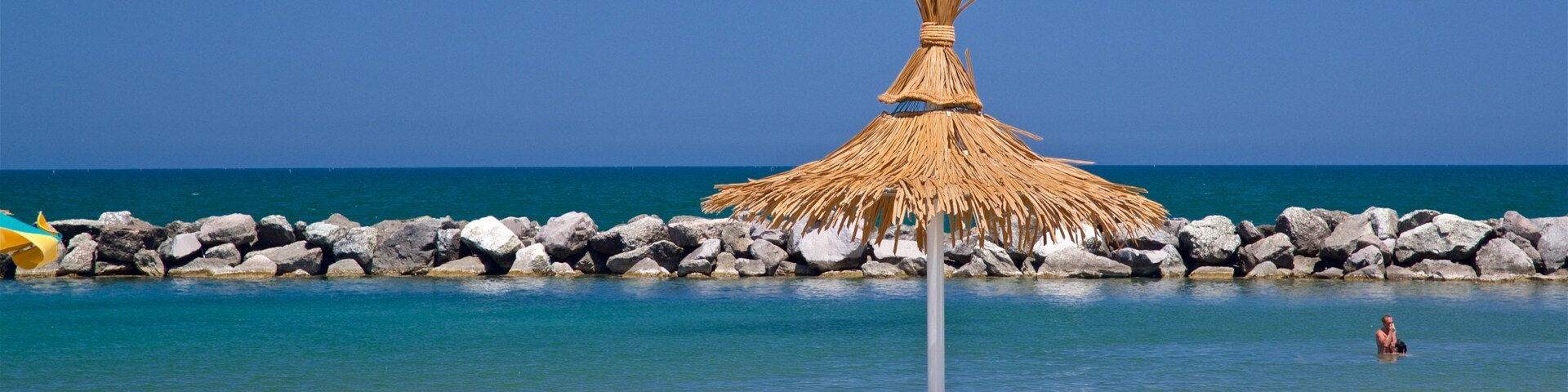 Uma praia com um guarda-sol de palha, um molhe rochoso e águas turquesa calmas.