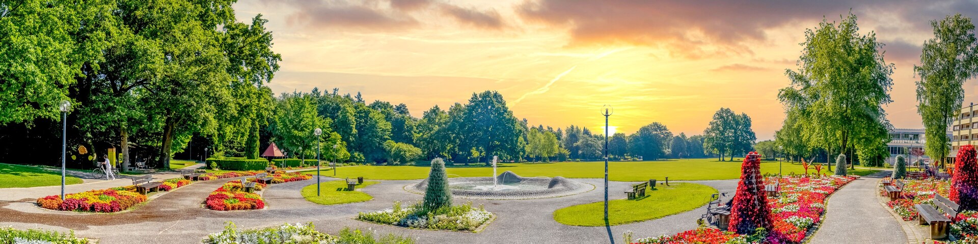 Um parque com canteiros de flores, um caminho sinuoso e um pôr do sol ao fundo.