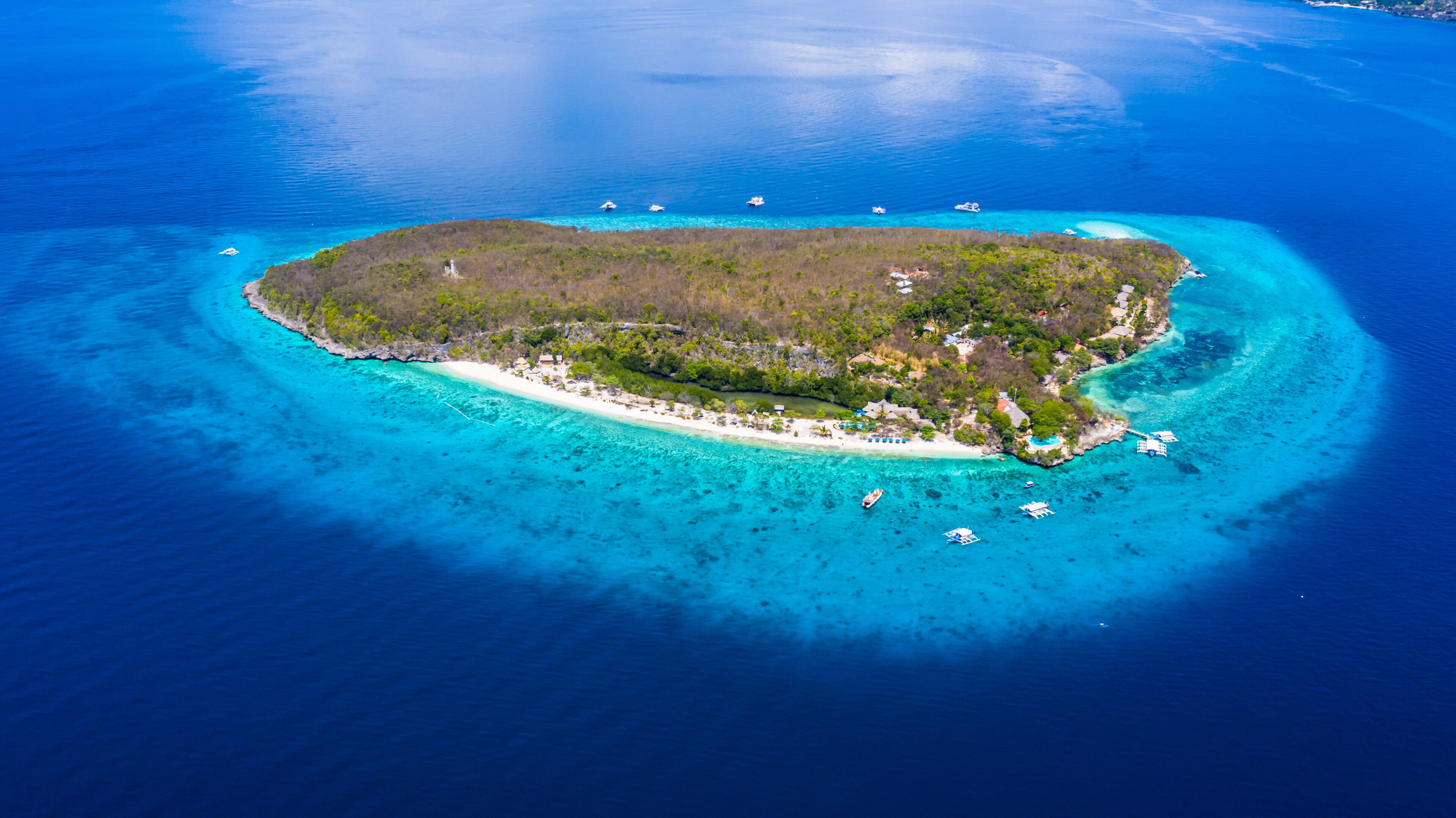 Vista aérea de uma pequena ilha com águas azul-turquesa cristalinas, rodeada por um recife de coral e algumas embarcações ancoradas perto da costa.