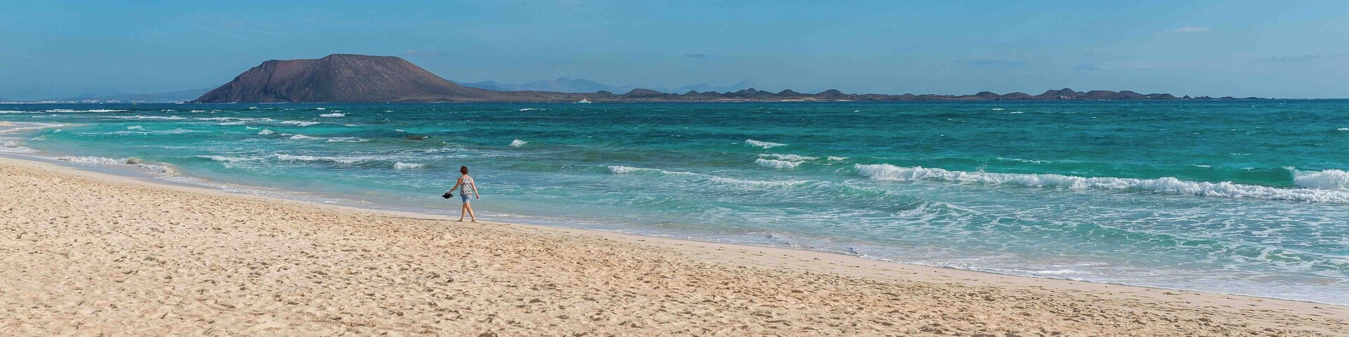 Uma praia arenosa com ondas suaves, céu claro e montanhas ao longe.