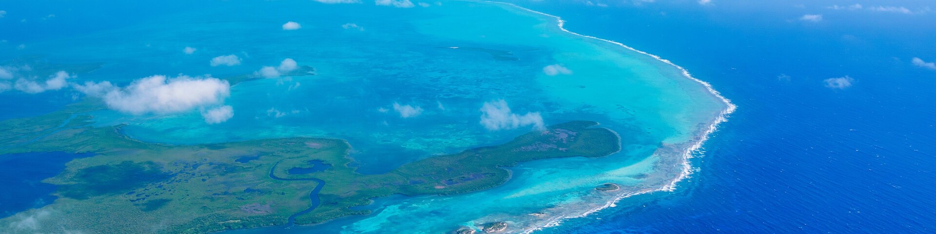 Vista aérea de uma região costeira com águas azul-claras, uma praia estreita e formações de nuvens dispersas.
