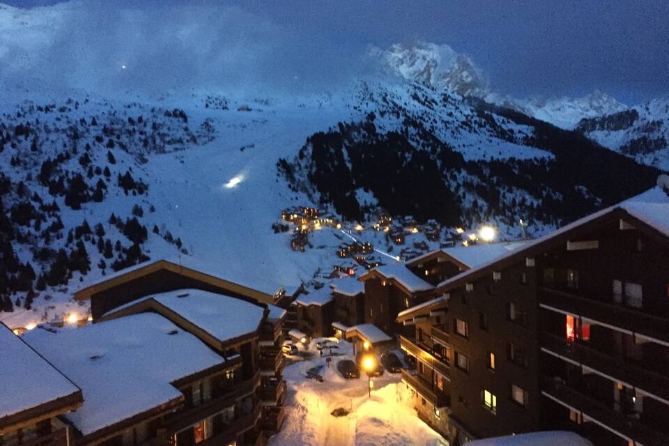 Uma cidade nas montanhas coberta de neve à noite, com edifícios iluminados e uma pista de esqui ao fundo.