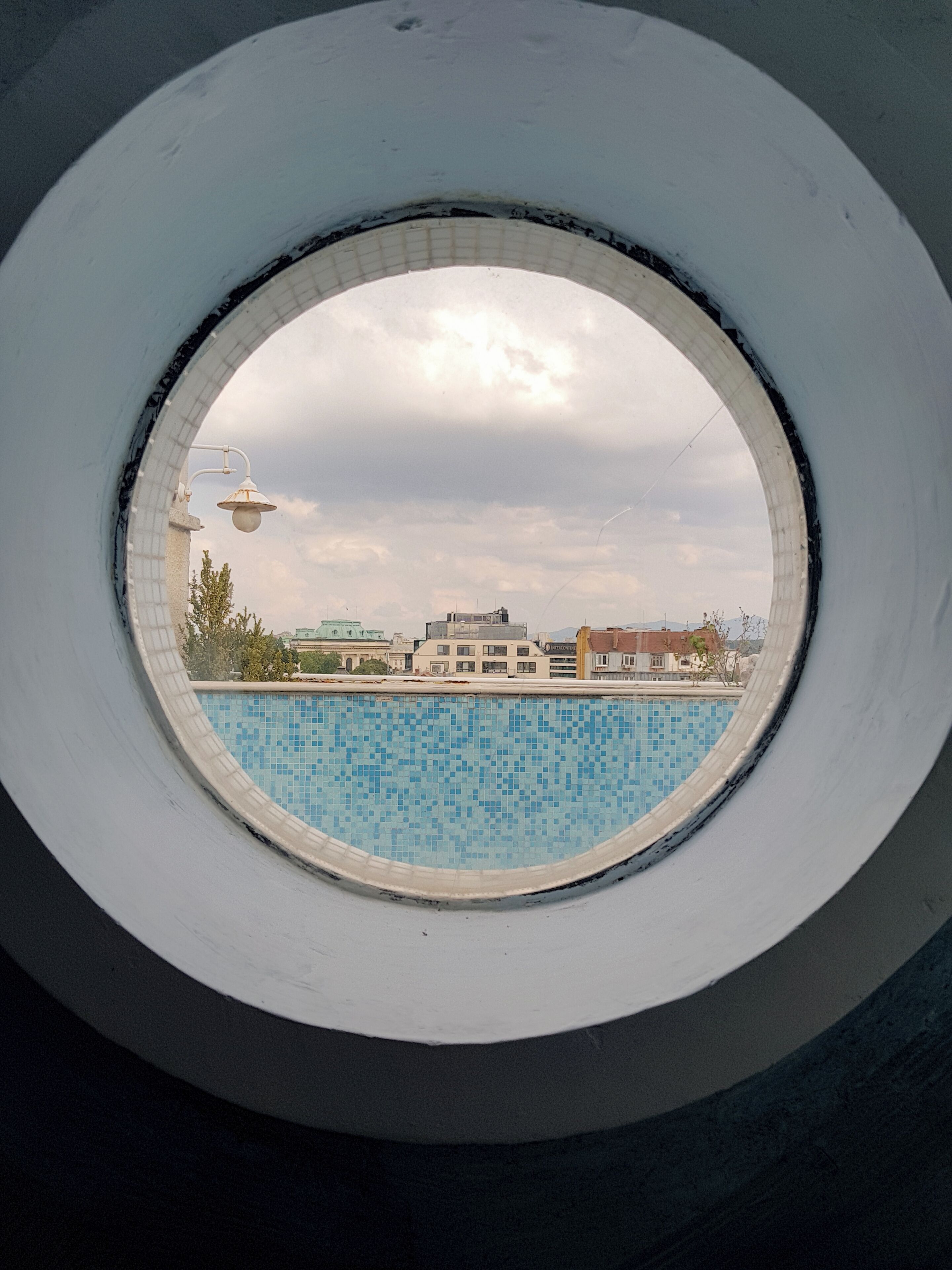 Janela redonda com vista para uma piscina e um deck revestido, com edifícios ao fundo.