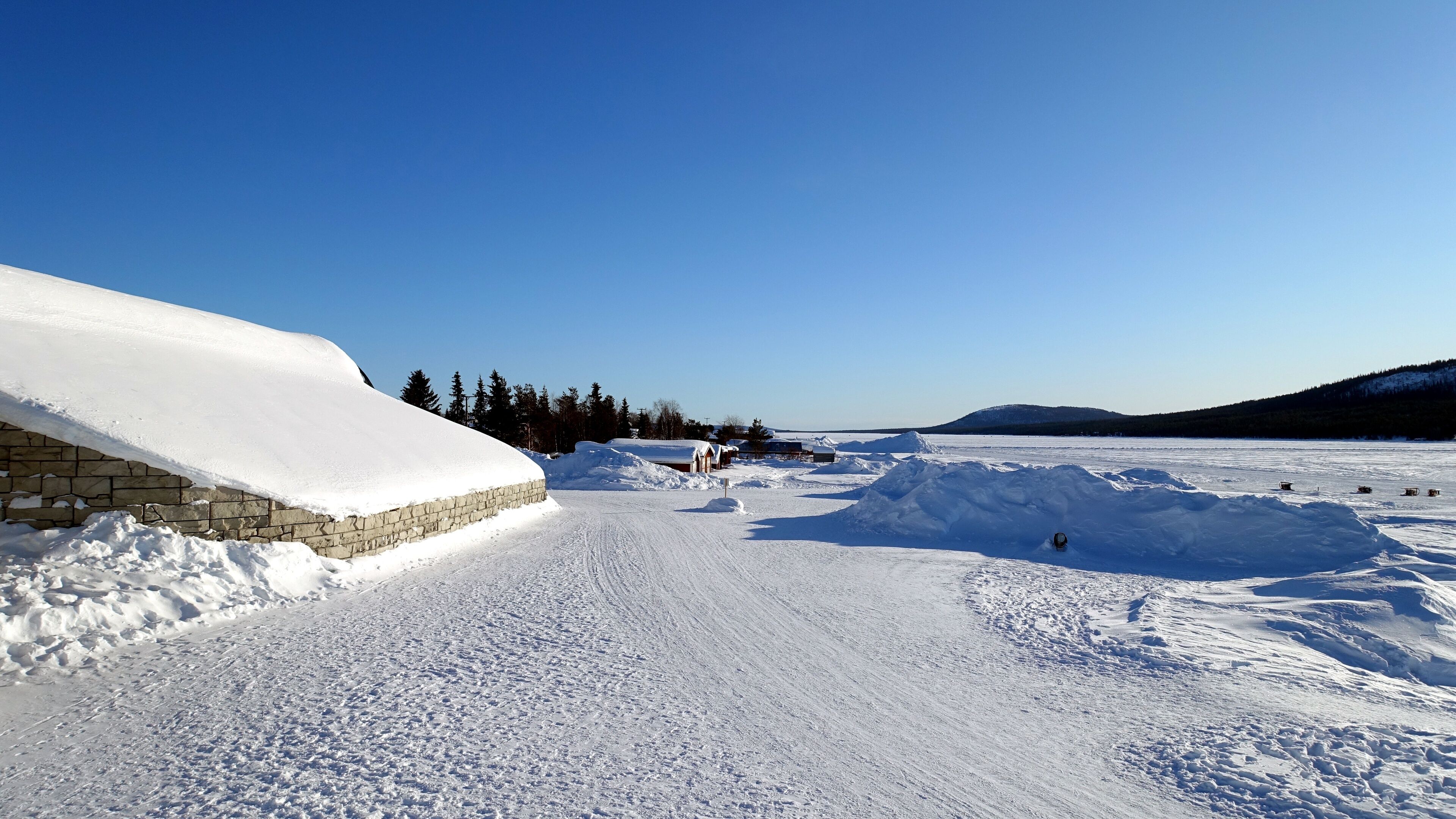 Paisagem coberta de neve com um muro de pedra, céu claro e montanhas ao longe.