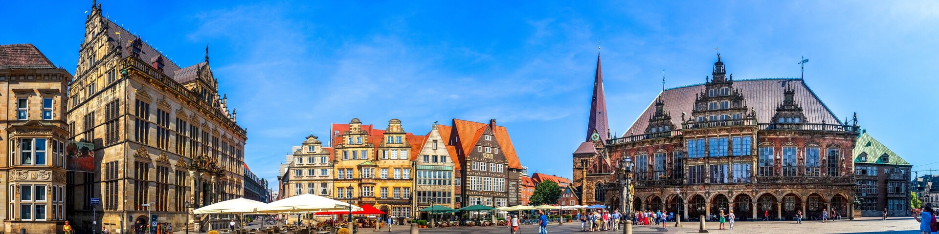 Uma praça histórica europeia com edifícios ricamente decorados, uma imponente torre de igreja e cafés ao ar livre.