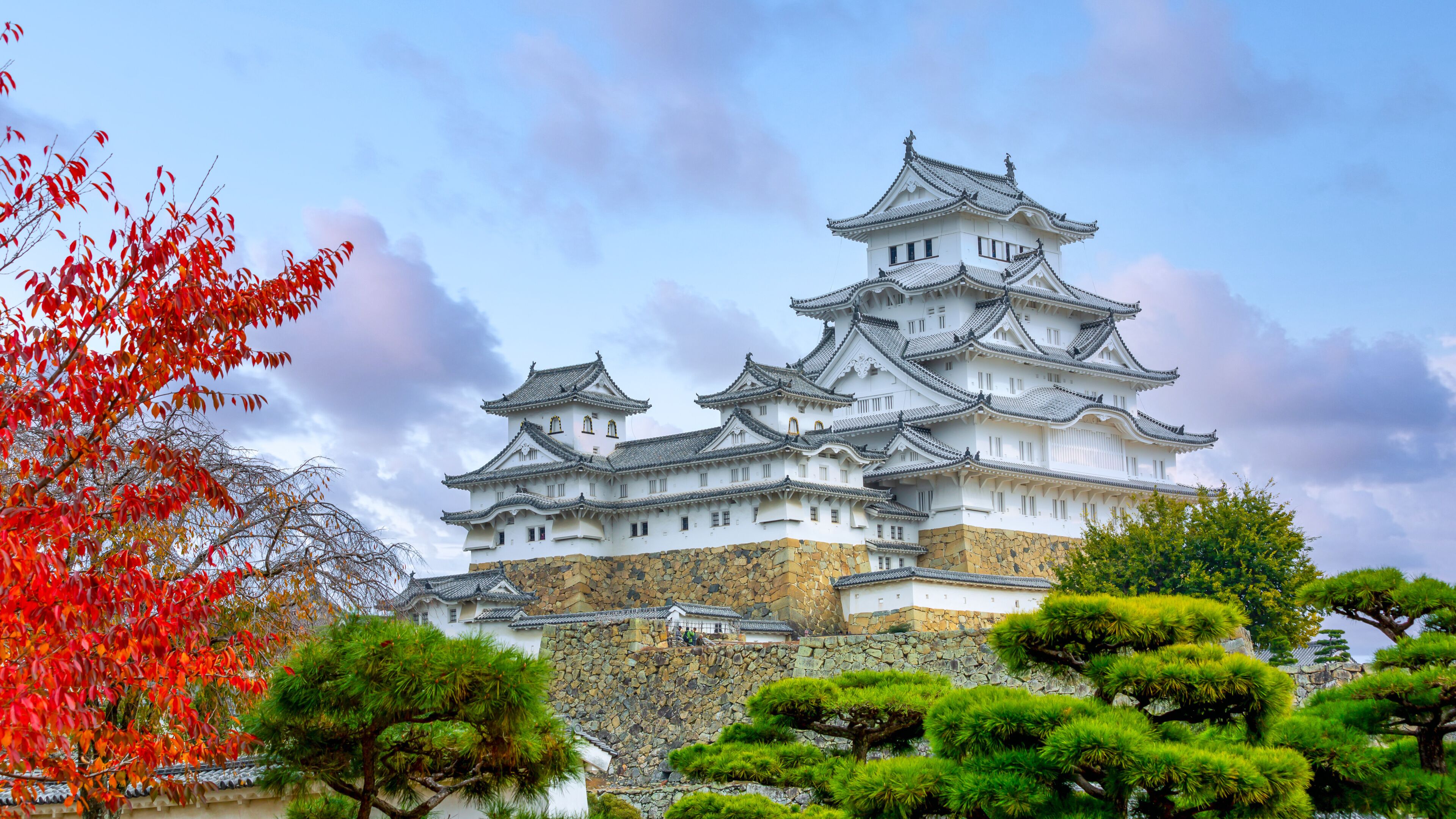 Um castelo japonês tradicional com vários níveis e base de pedra, rodeado por vegetação exuberante e árvores de cores outonais.