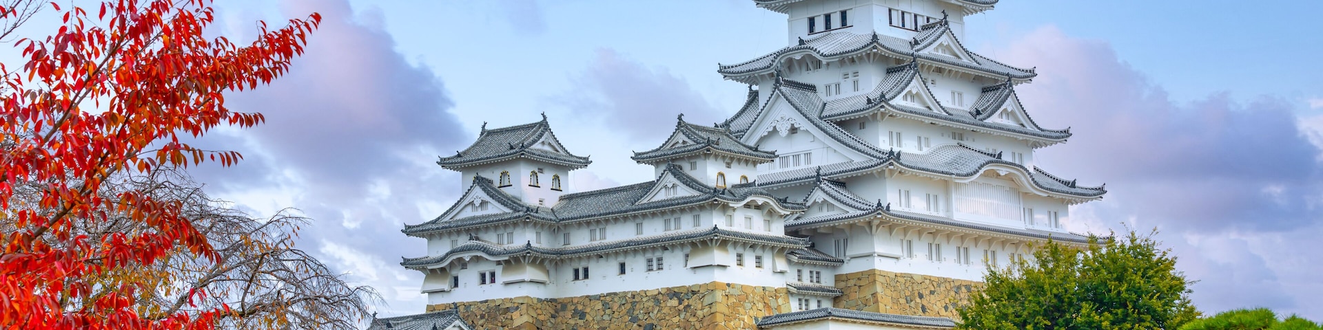 Um castelo japonês tradicional com vários níveis e base de pedra, rodeado por vegetação exuberante e árvores de cores outonais.