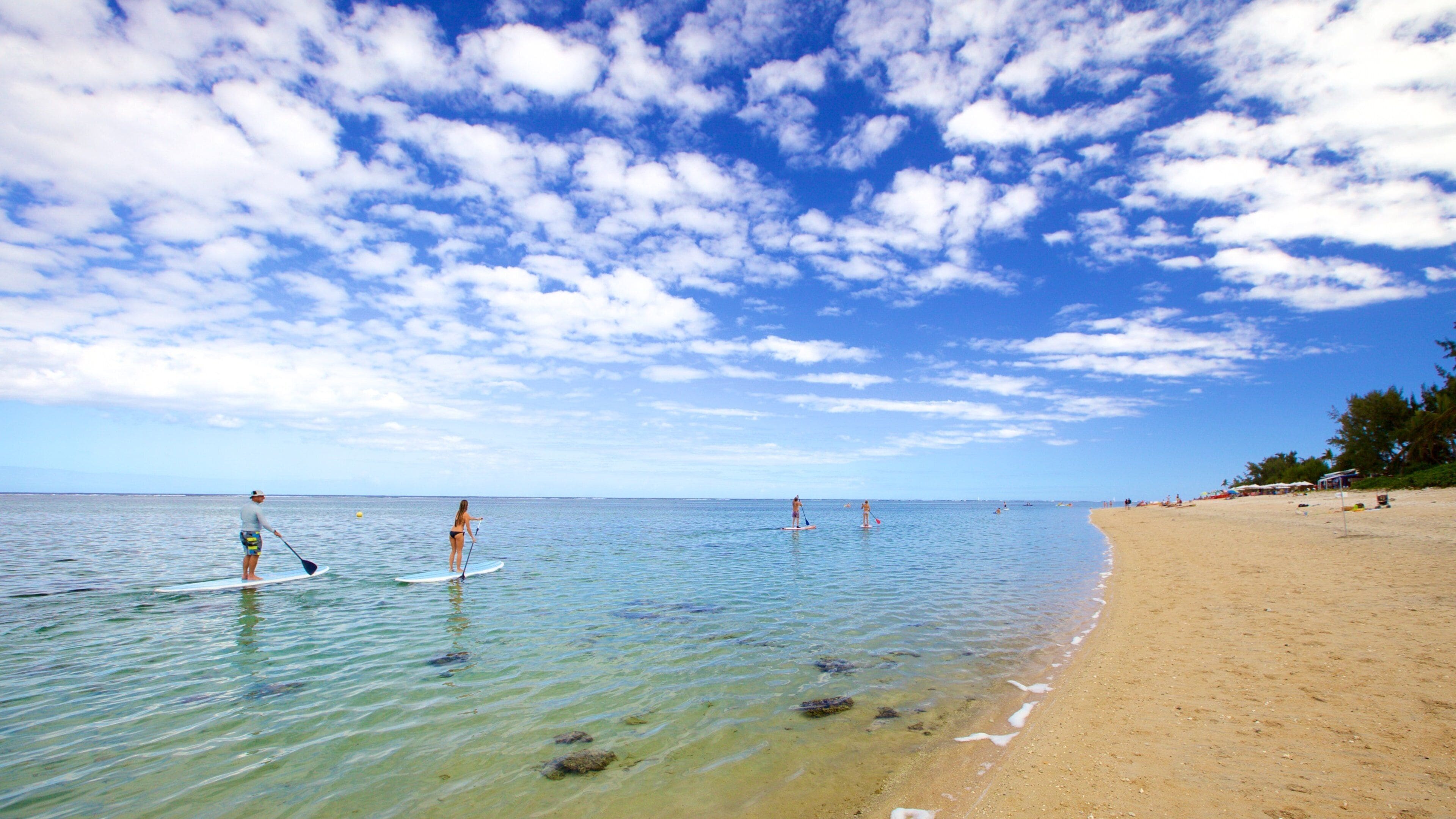 Uma praia com águas claras, pessoas praticando paddleboard e uma faixa de areia sob um céu parcialmente nublado.