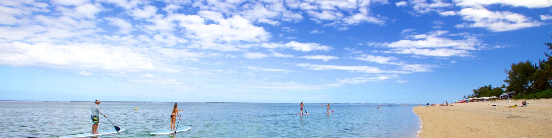 Uma praia com águas claras, pessoas praticando paddleboard e uma faixa de areia sob um céu parcialmente nublado.
