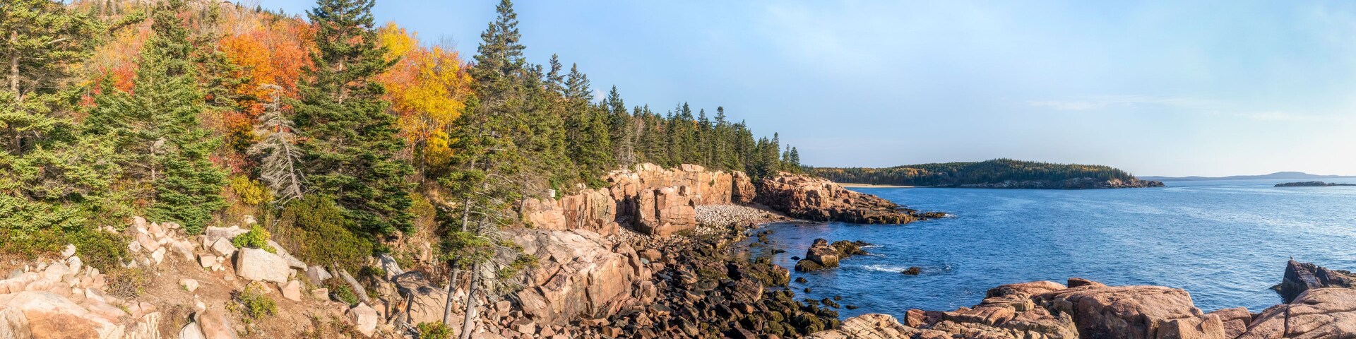 Paisagem costeira com falésias rochosas, árvores em tons de outono e mar calmo.
