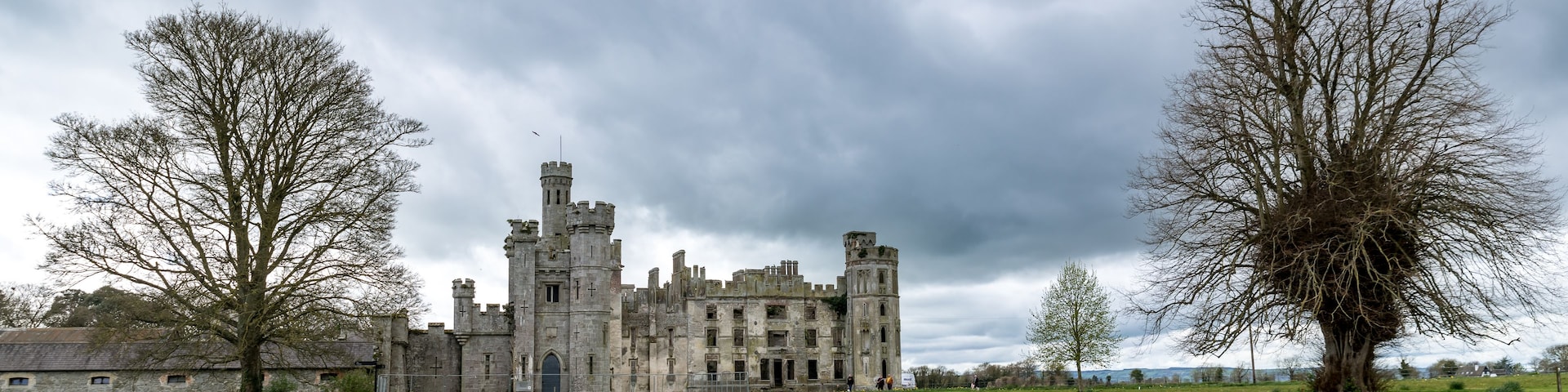 Um castelo histórico com várias torres e ameias, cercado por um campo gramado e árvores sem folhas sob um céu nublado.
