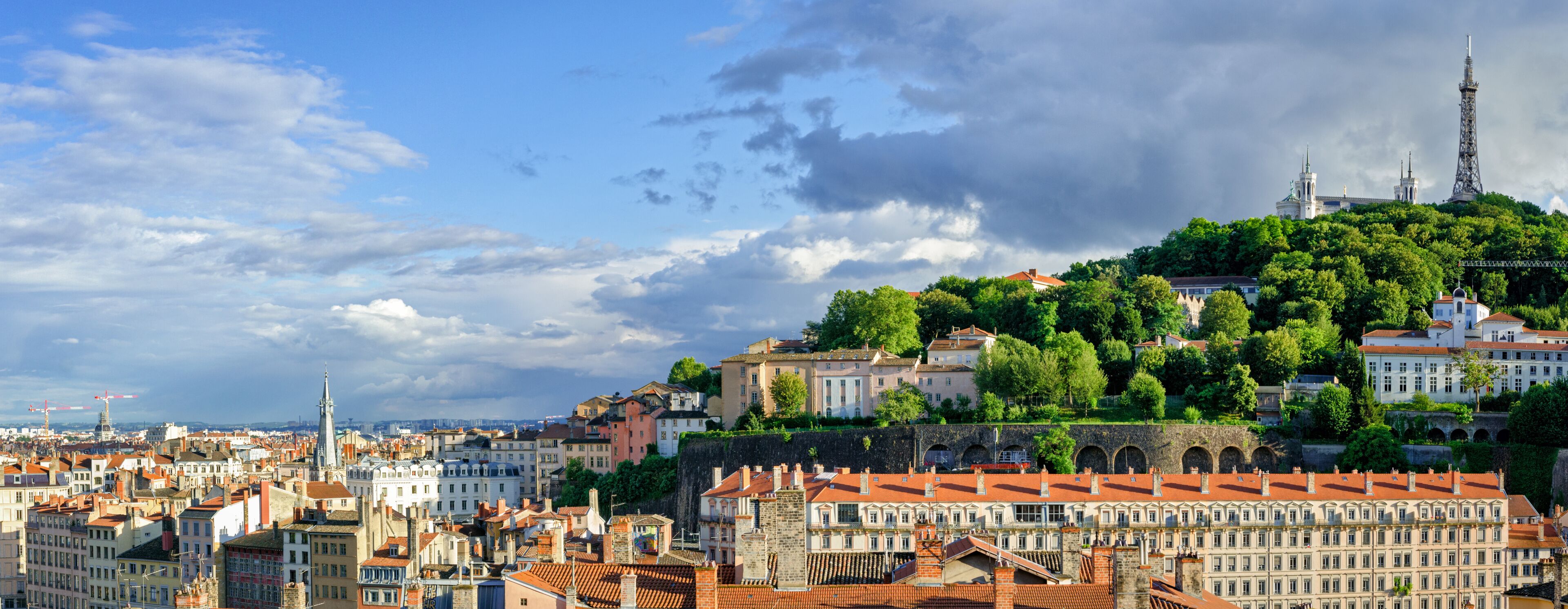 Paisagem urbana com edifícios históricos, uma torre imponente e vegetação exuberante numa colina.