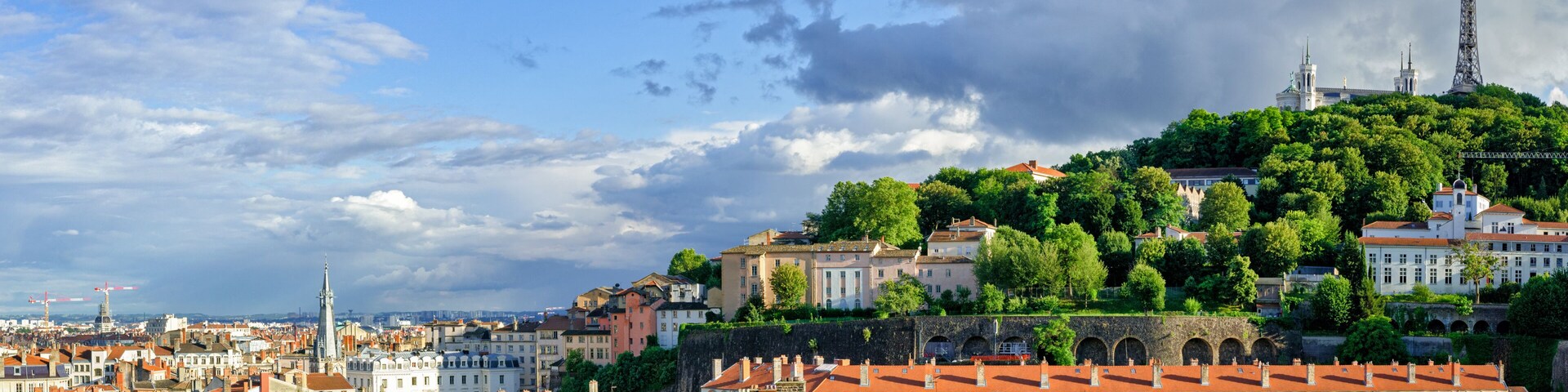 Paisagem urbana com edifícios históricos, uma torre imponente e vegetação exuberante numa colina.