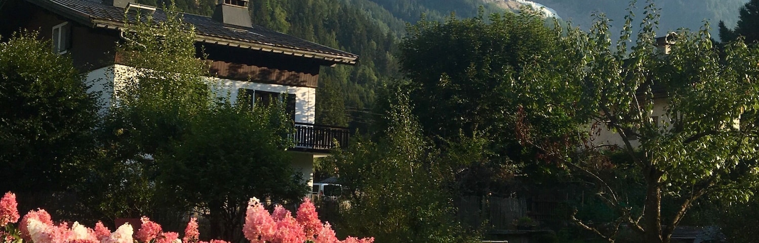Paisagem de montanha com picos nevados, uma casa e flores rosa em primeiro plano.