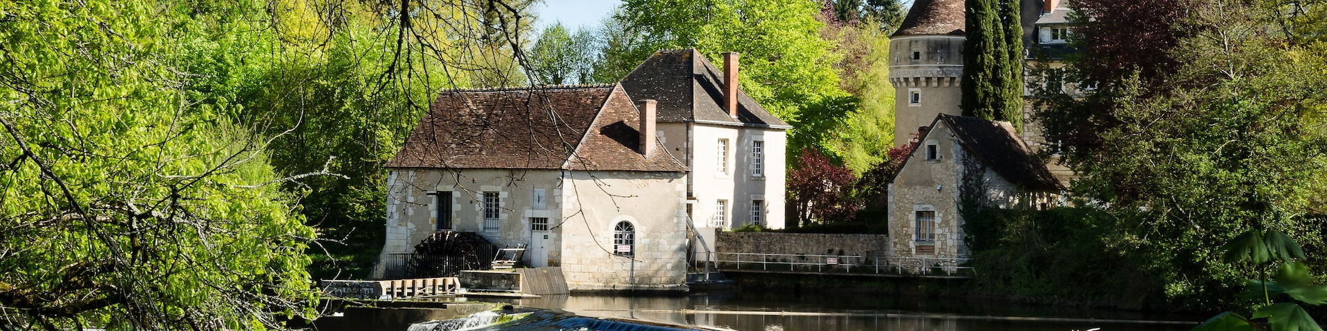 Um edifício histórico com uma roda de moinho, cercado por vegetação exuberante e um rio calmo.