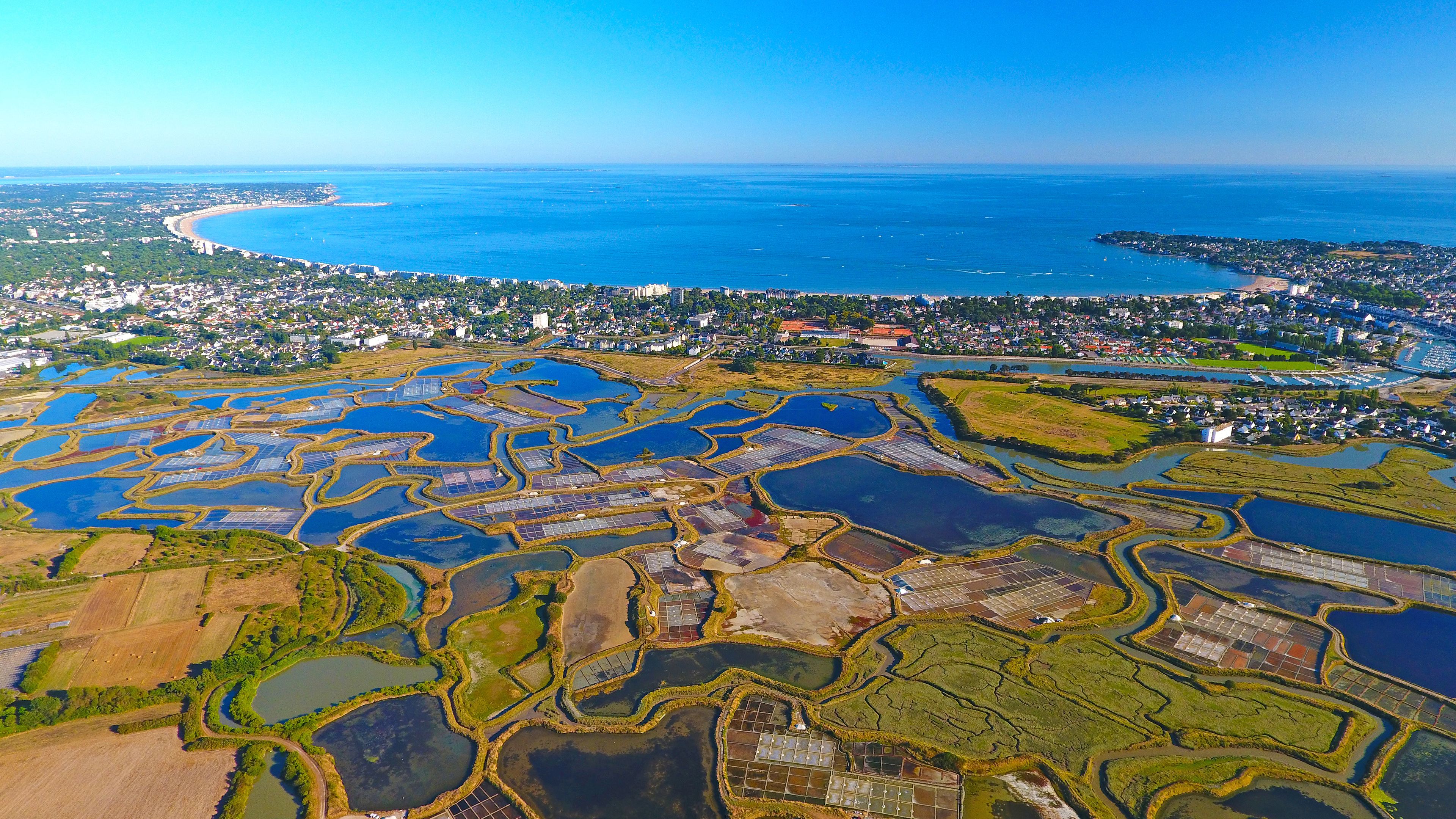 Vista aérea de uma área costeira com campos em socalcos, uma praia e um horizonte urbano distante.