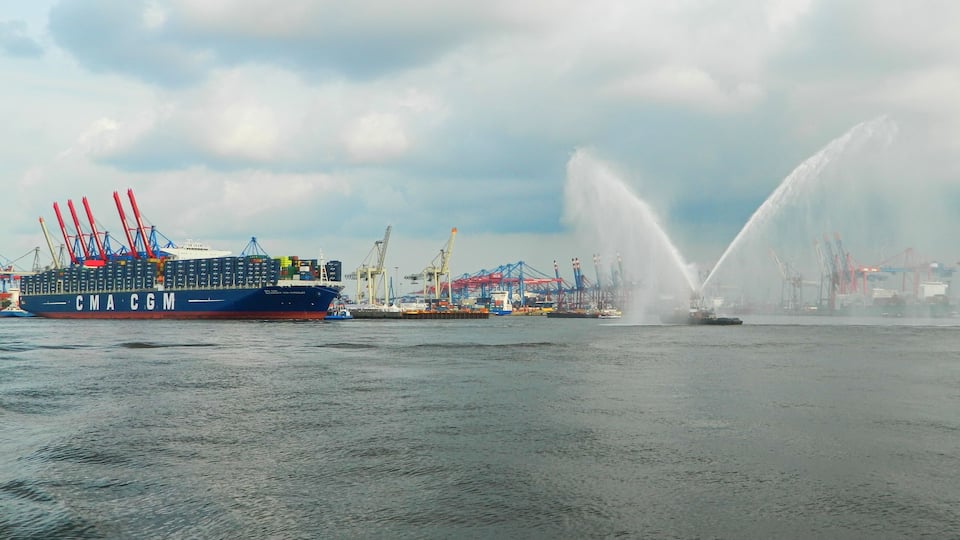 Um navio de carga com as letras "CMA CGM" na lateral, atracado em um porto com guindastes e uma fonte ao fundo.